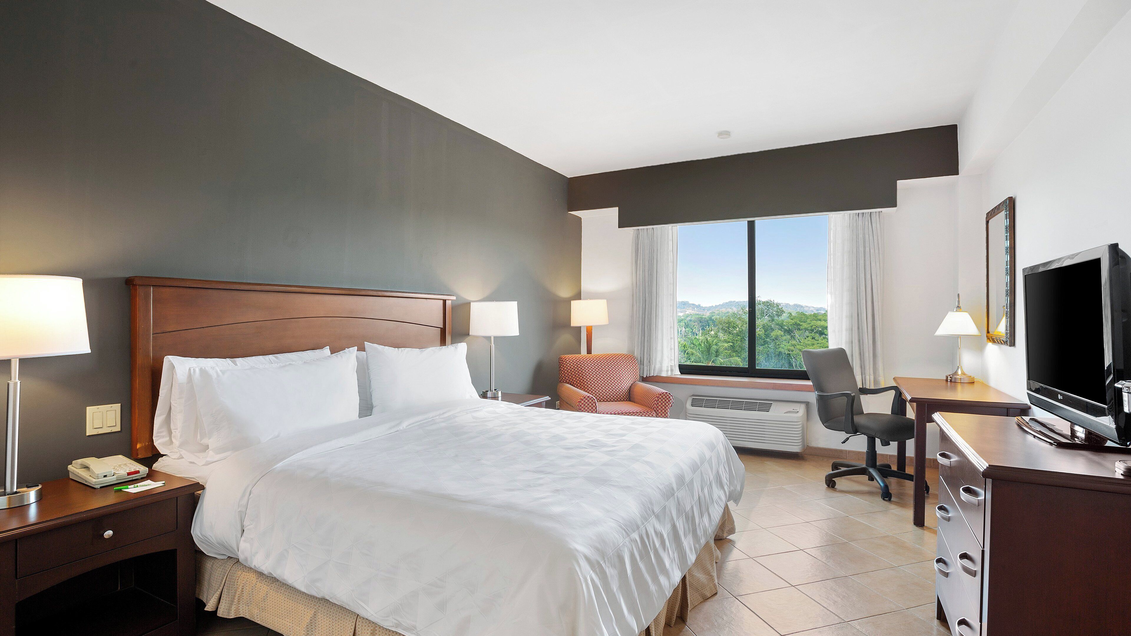 Quarto de hotel com uma cama grande, duas mesas de cabeceira com luminárias, uma escrivaninha com televisão, uma cadeira e uma janela com vista para o verde.