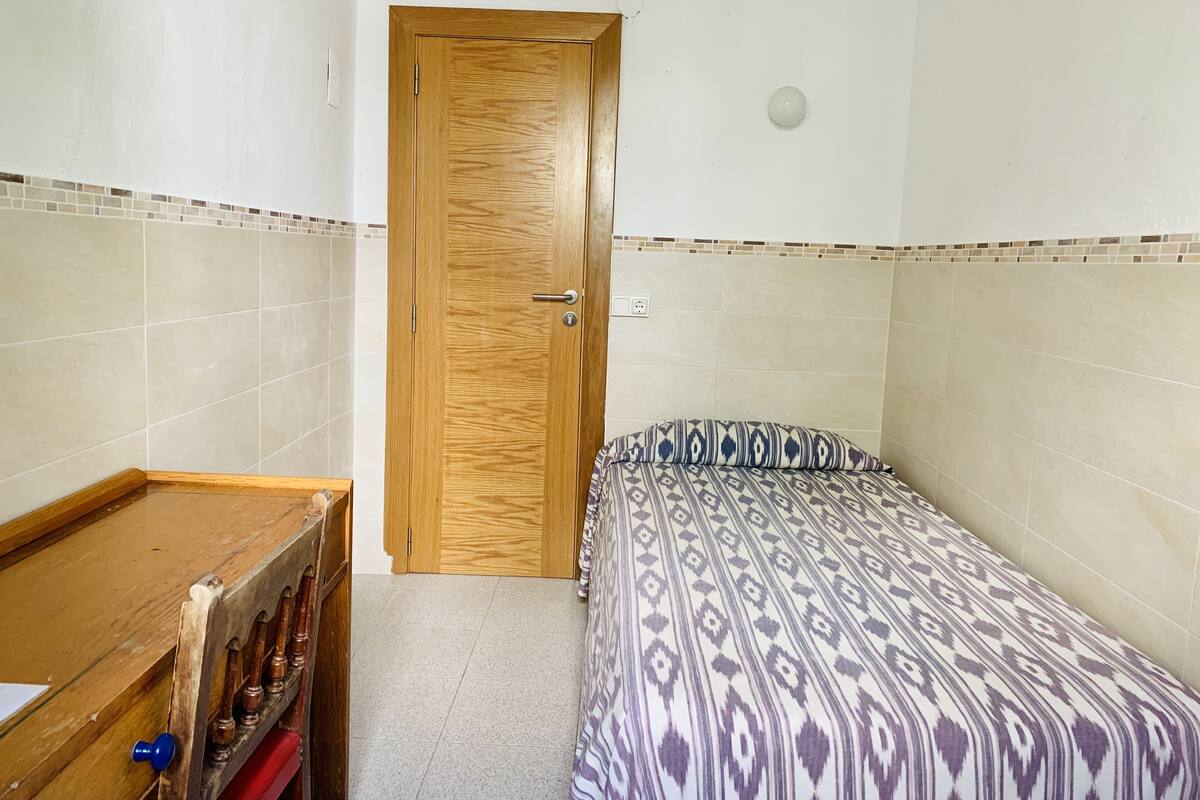 Um quarto pequeno e limpo, com porta de madeira, cama com roupa de cama estampada e mesa de madeira com um tecido vermelho.