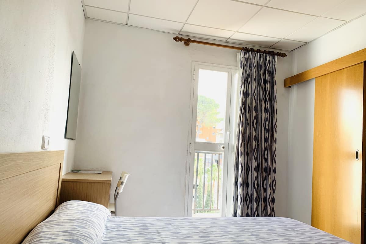 Quarto com cama, cabeceira de madeira, mesa de cabeceira, janela com cortina e porta.