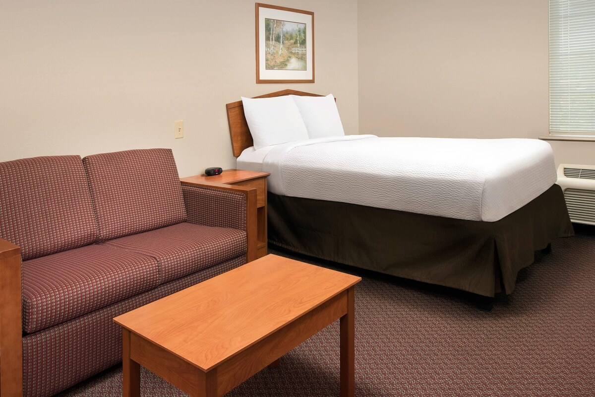 Quarto de hotel com cama, sofá, mesa pequena e relógio fixado na parede.