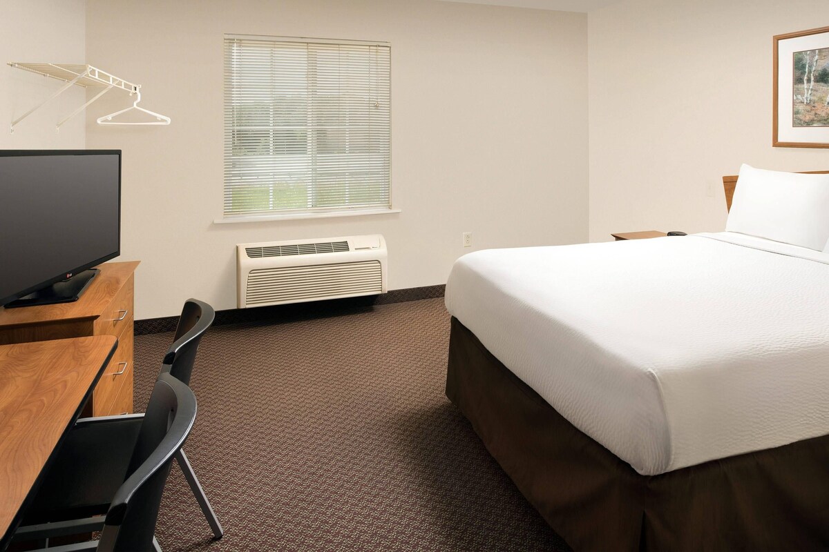 Quarto de hotel com cama, escrivaninha com televisão, cadeira, janela com persianas e aquecedor fixado na parede.