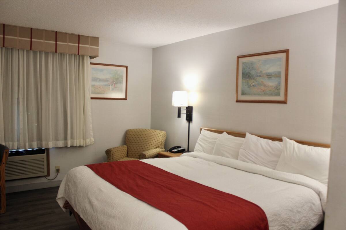 Quarto de hotel com uma cama grande, uma cadeira, uma escrivaninha e duas pinturas nas paredes.