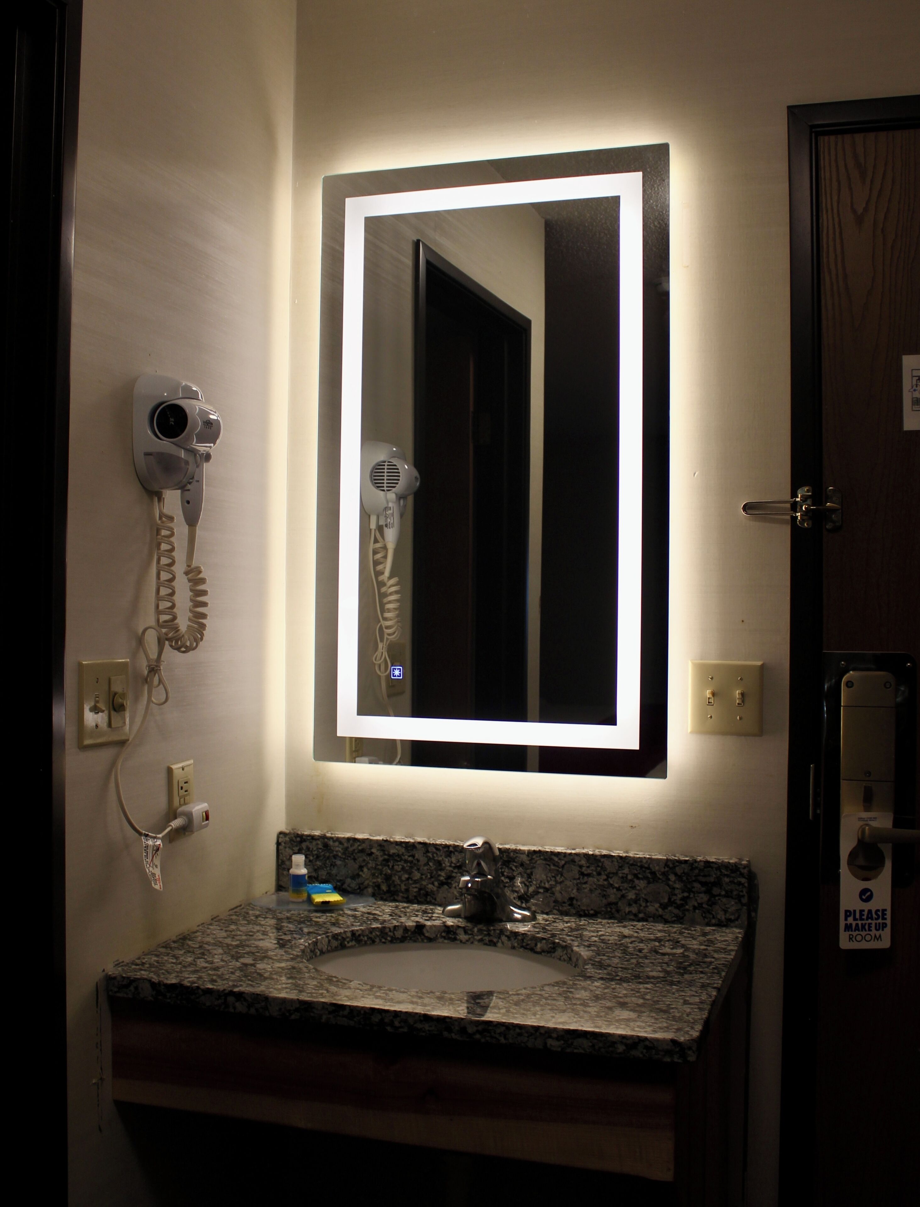 Casa de banho com bancada de granito, espelho iluminado, secador de cabelo e porta com a placa 'Make Up Room'.