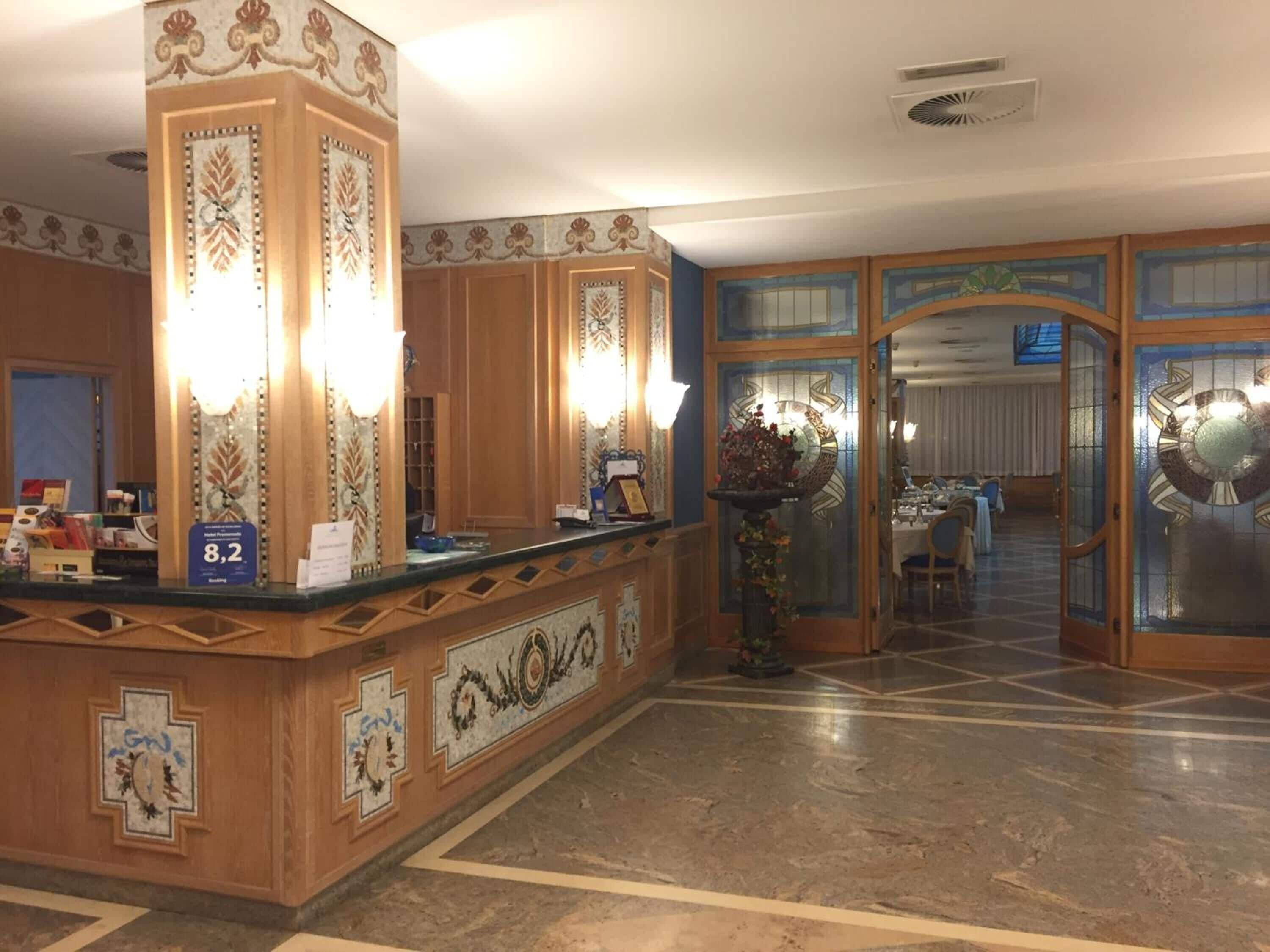 Uma área de recepção de hotel com piso de mármore, um balcão de atendimento com placa azul e um espelho decorativo grande.