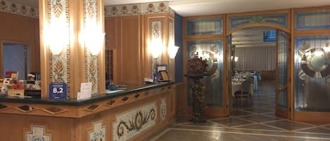 Uma área de recepção de hotel com piso de mármore, um balcão de atendimento com placa azul e um espelho decorativo grande.