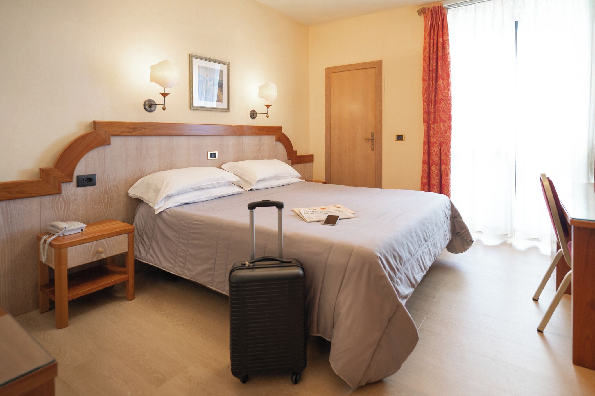 Quarto de hotel com uma cama, mesa de cabeceira de madeira com um telefone, uma mala e uma escrivaninha com uma cadeira.