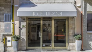 A entrada do Hotel Lupori, com fachada de vidro transparente, uma placa acima da entrada e duas plantas em vasos de cada lado.