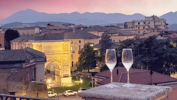 Vista do pôr do sol de uma cidade com montanhas ao fundo, um edifício histórico com um arco e dois copos de vinho vazios sobre uma mesa.