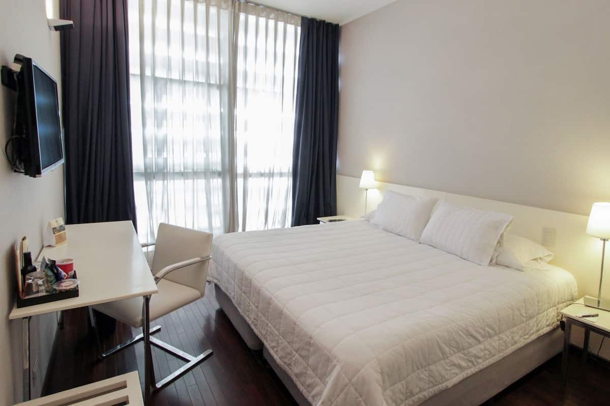 Quarto de hotel com cama, escrivaninha com cadeira, televisão na parede e janela com cortinas.