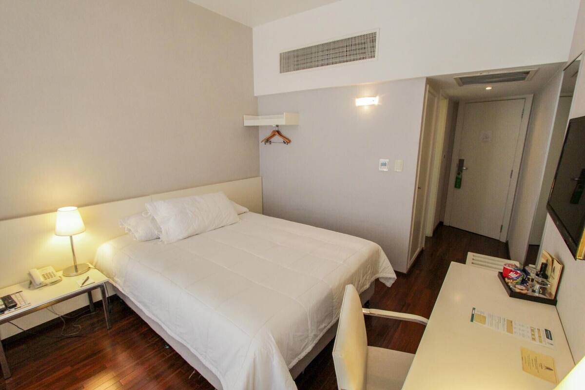 Quarto de hotel com cama, mesa de cabeceira com abajur, escrivaninha com cadeira e televisão.