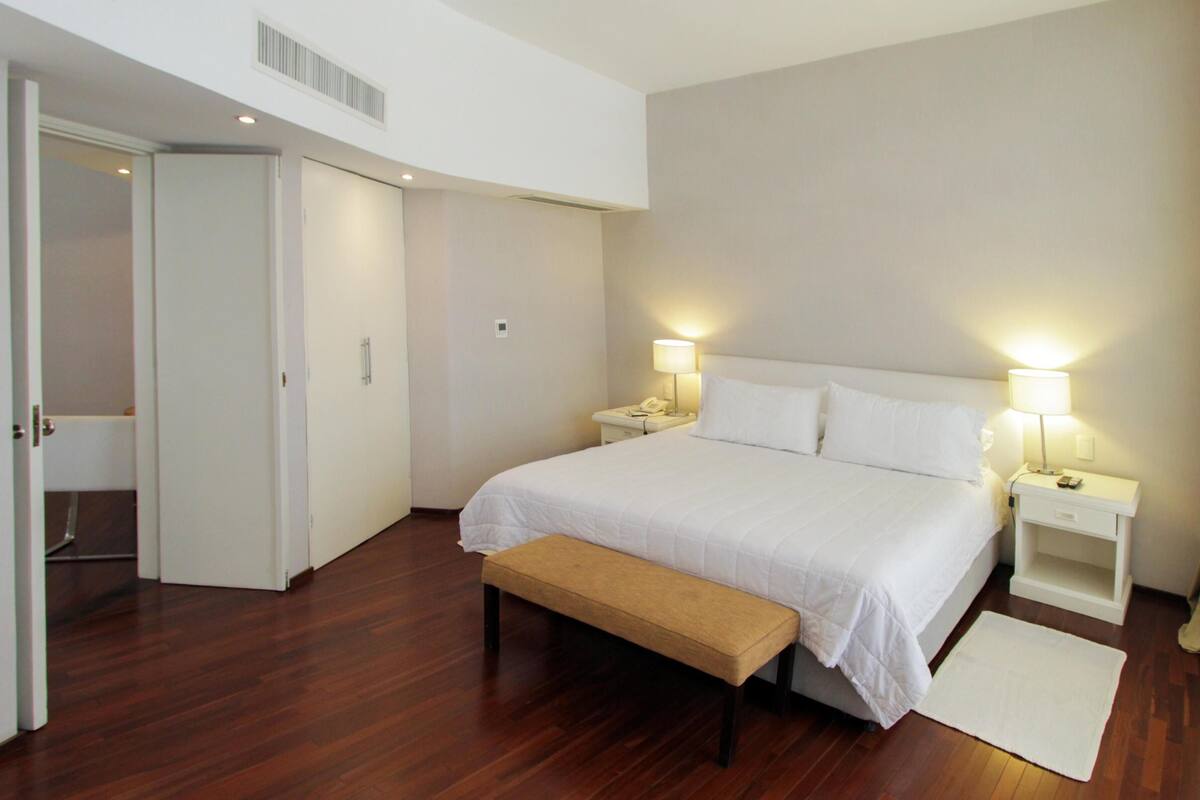 Quarto de hotel com uma cama grande, abajures, um banquinho e um tapete branco.
