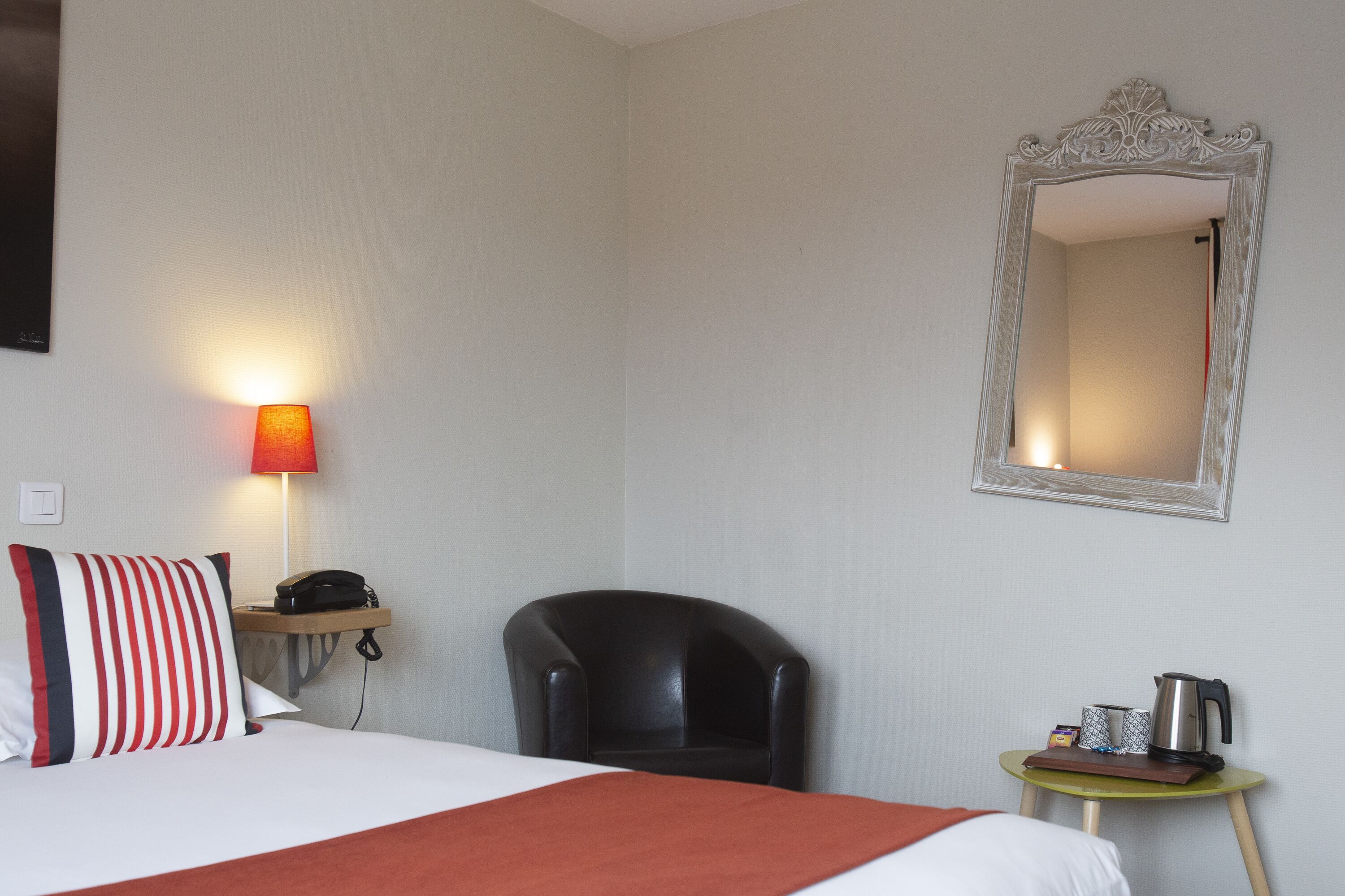 Quarto de hotel com cama, uma cadeira, uma mesinha com chaleira, abajur e espelho.
