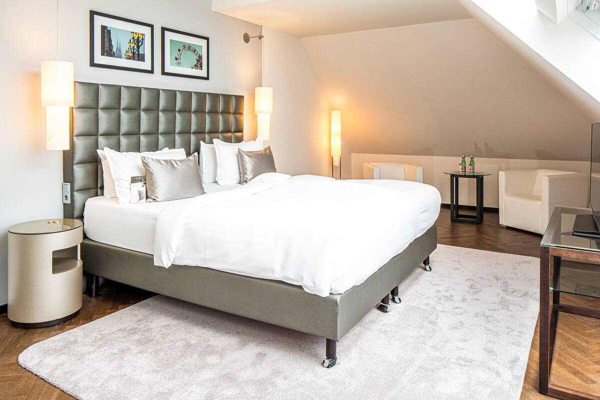 Quarto de hotel moderno com uma cama grande, duas luminárias de cabeceira, uma mesinha redonda, uma mesinha lateral pequena, um sofá e uma televisão.