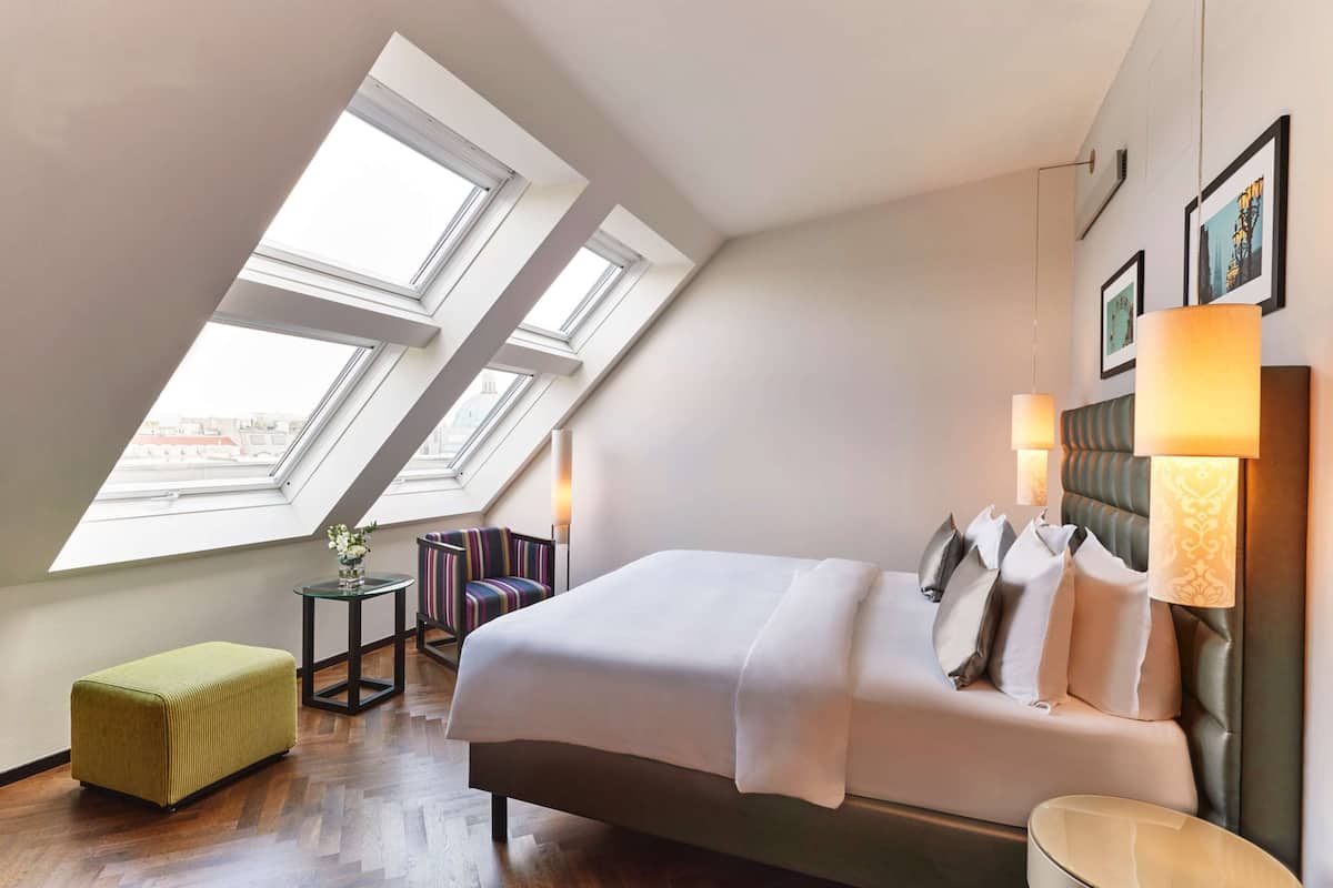 Quarto de hotel moderno com uma cama grande, janelas tipo claraboia, uma mesinha lateral, um banquinho verde e uma cadeira.