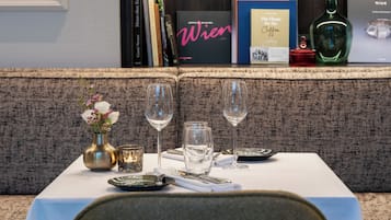 Uma mesa de jantar posta com taças de vinho, pratos e talheres, com uma estante de livros ao fundo.