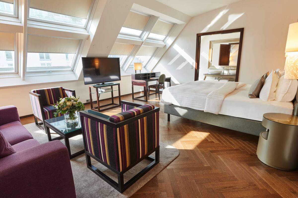 Quarto de hotel moderno com cama, escrivaninha, televisão e área de estar com cadeiras listradas.