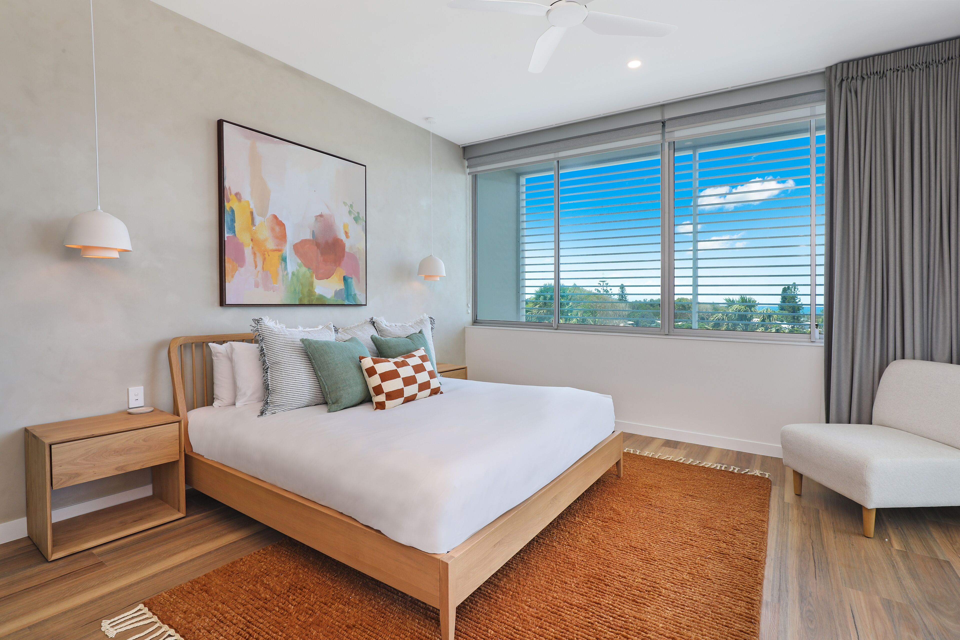 Quarto moderno com uma cama grande, mesas de cabeceira, ventoinha no teto, janela com persianas e vista para o verde lá fora.