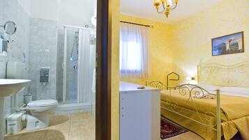 Casa de banho com chuveiro, sanita e lavatório. Inclui um espelho. O quarto tem uma cama com colcha amarela e uma mesa de cabeceira com candeeiro.