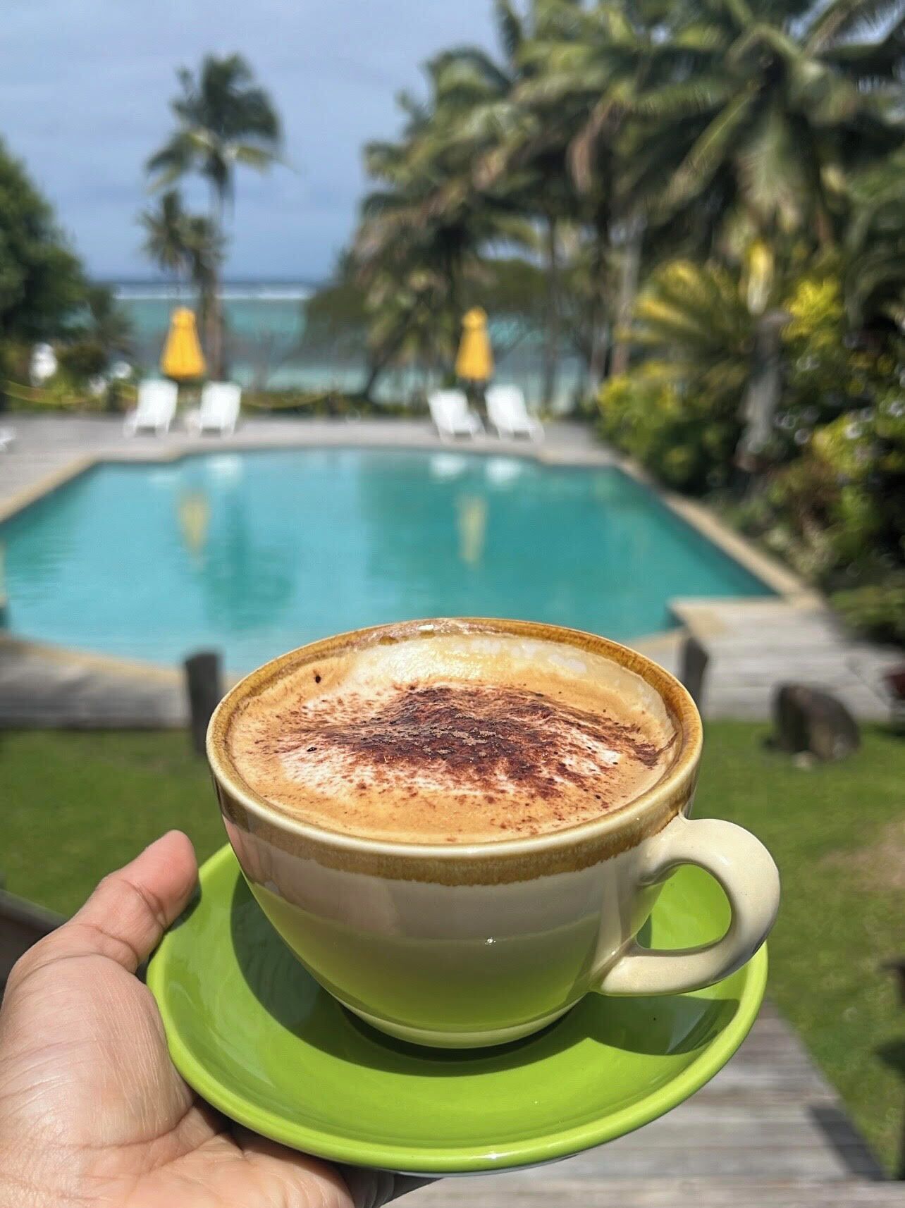 Uma xícara de café com espuma, segurada na mão, com uma piscina e palmeiras ao fundo.