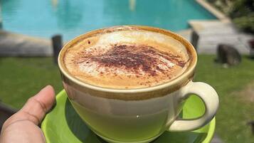 Uma xícara de café com espuma, segurada na mão, com uma piscina e palmeiras ao fundo.