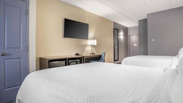 Quarto de hotel com duas camas, uma escrivaninha com candeeiro, televisão de ecrã plano na parede e uma porta.