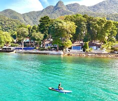 Uma área costeira com águas turquesa cristalinas, uma pessoa praticando paddleboard e casas com vista para as montanhas ao fundo.