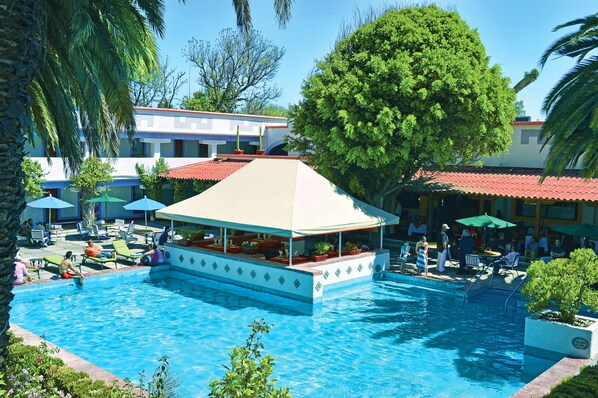 Piscina com espreguiçadeiras e guarda-sóis, cercada por palmeiras e um edifício com telhado vermelho.