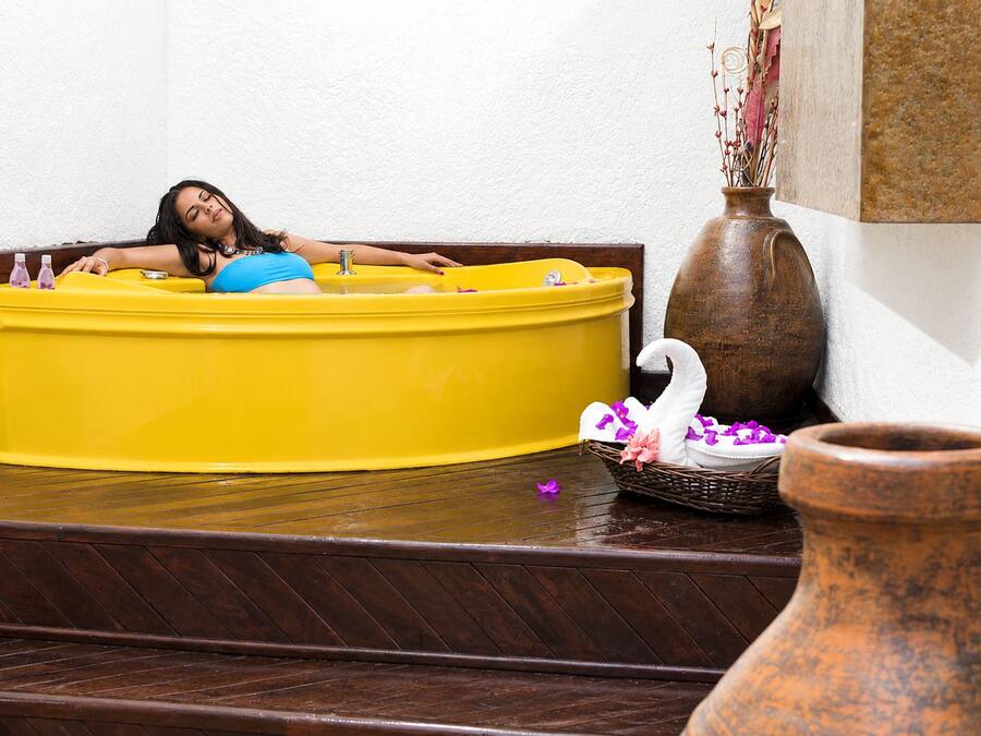 Uma pessoa relaxando em uma banheira amarela com tapete de banho em formato de cisne e um vaso com flores secas.