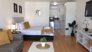 Quarto de hotel moderno com cama, sofá, uma pequena mesa com planta, área de cozinha e televisão fixada na parede.