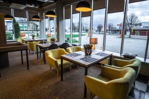 Restaurante com cadeiras amarelas, mesas montadas e amplas janelas com vista para a rua.