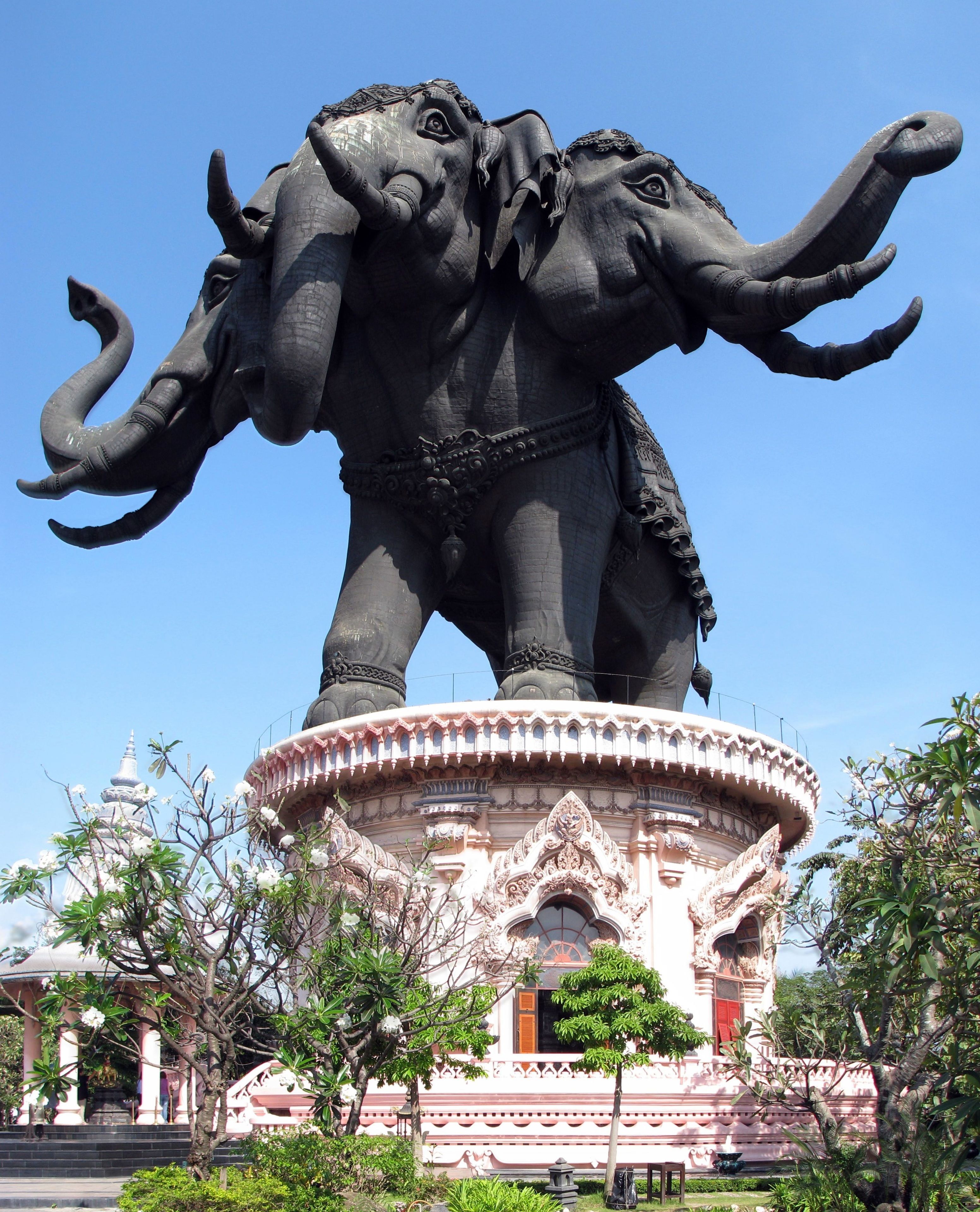 Uma grande estátua de elefante sobre um pedestal circular com esculturas detalhadas, rodeada por árvores e um templo ao fundo.
