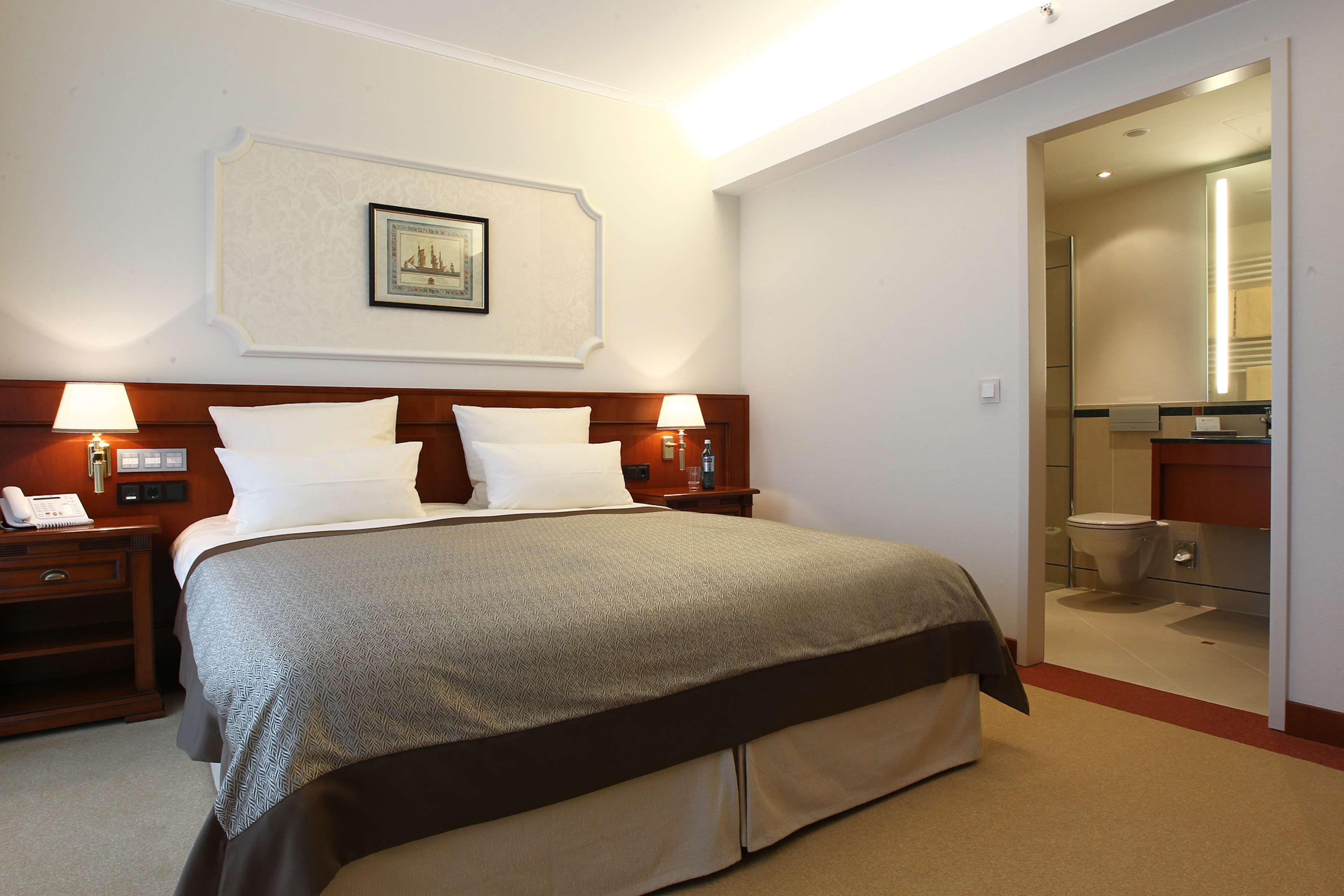 Quarto de hotel com uma cama grande, duas mesas de cabeceira com luminárias, um quadro na parede e um banheiro visível por uma porta aberta.