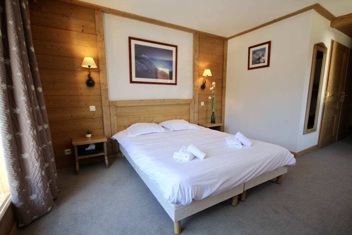 Cama bem arrumada com lençóis e toalhas brancos, cabeceira de madeira e duas luminárias fixadas na parede.