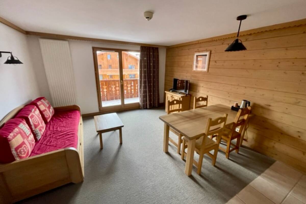 Quarto com piso de madeira, sofá, mesa e cadeiras. Janela com vista para prédios.