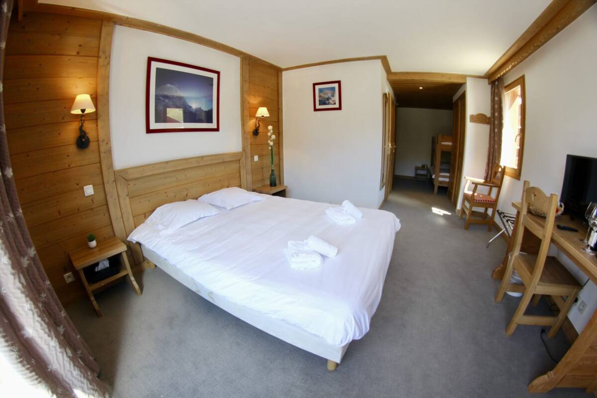 Quarto de hotel com uma cama grande, paredes de madeira, duas luminárias laterais, uma pequena mesa com uma planta e uma televisão em um suporte de madeira.