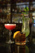 Um bar com um coquetel em um copo, uma jarra com um ramo de hortelã e um fundo desfocado de prateleiras com garrafas.