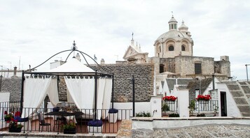 Um edifício de pedra tradicional com cúpula e torre de sino, um coreto branco com estrutura de metal e plantas em vasos.