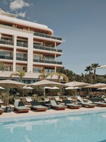 Um hotel com área de piscina, espreguiçadeiras, guarda-sóis e um edifício de vários andares com varandas.