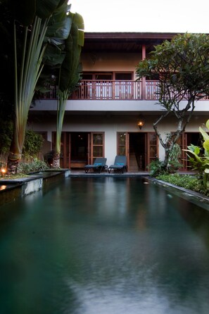 Um edifício de dois andares com piscina, espreguiçadeiras e plantas tropicais.