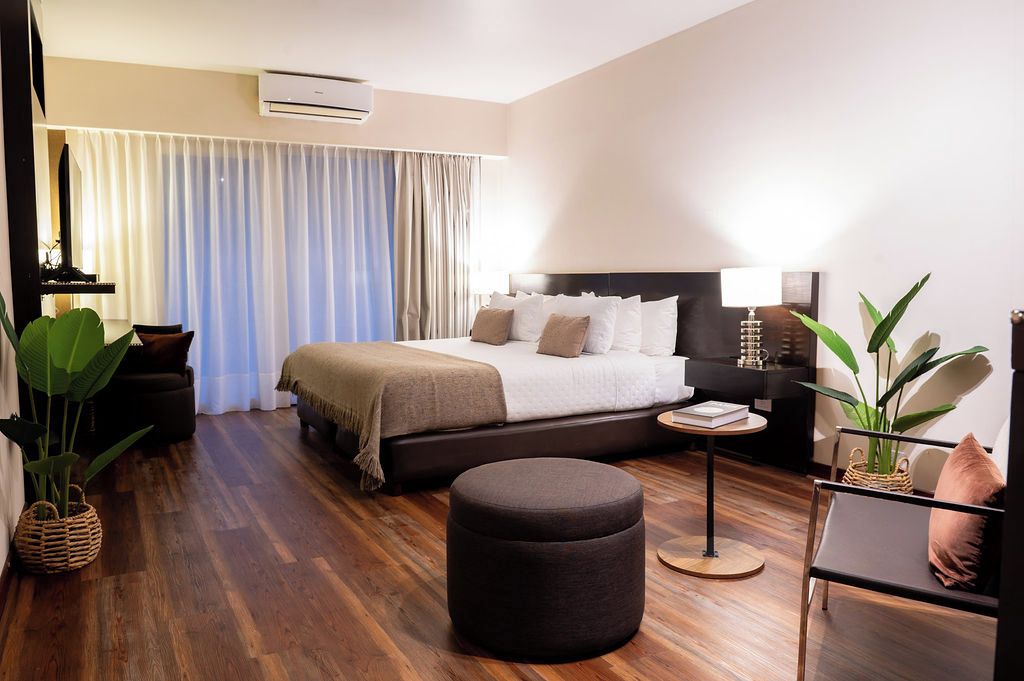 Quarto de hotel moderno com uma cama grande, uma mesa de cabeceira, uma pequena mesa redonda e um espaço de estar com uma poltrona.