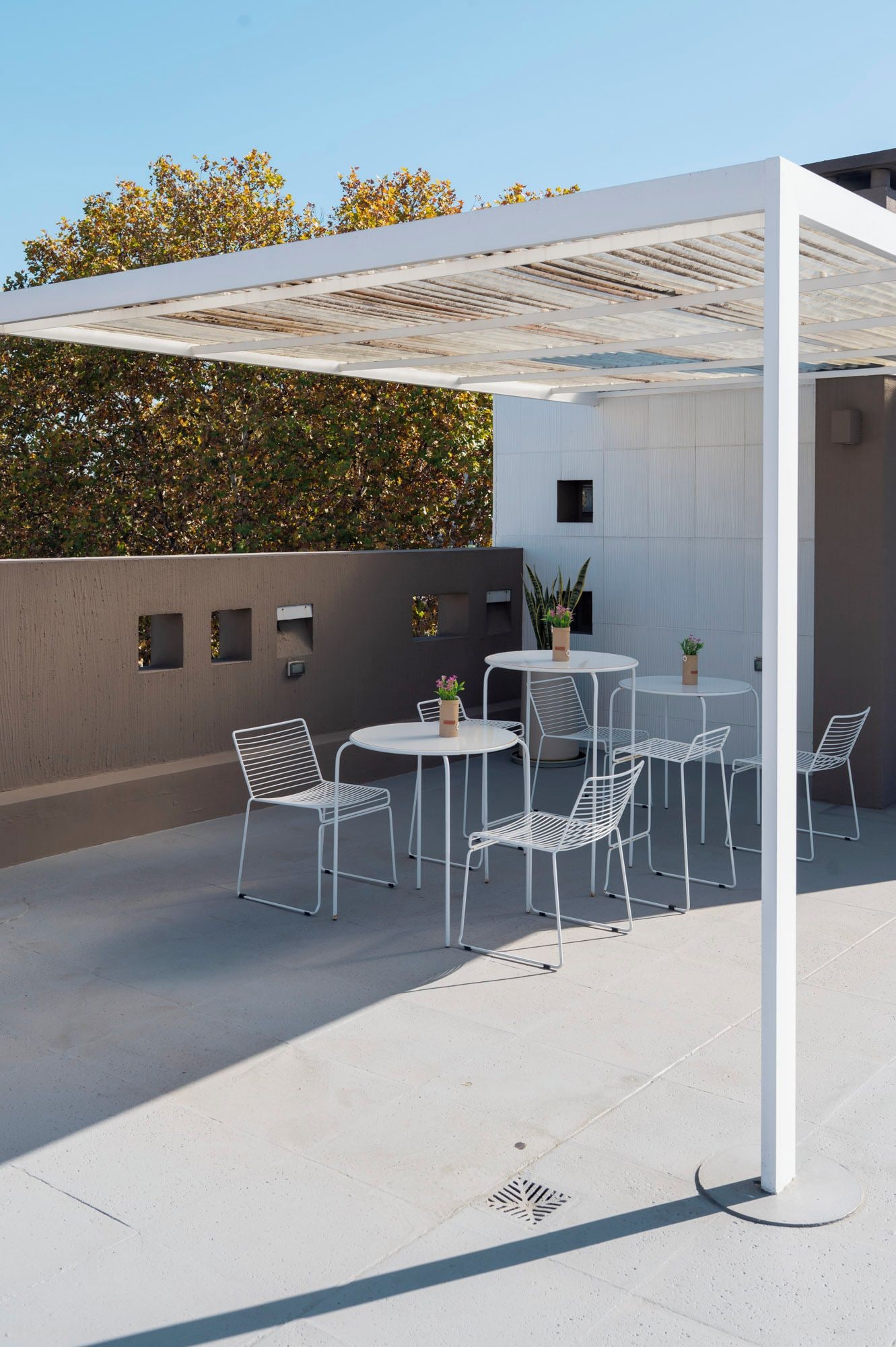 Um pátio externo com cadeiras e mesas de metal branco, uma pérgola e uma parede com janelas pequenas.
