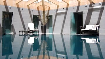 Piscina interior moderna com água cristalina, vigas de madeira e duas cadeiras de descanso.