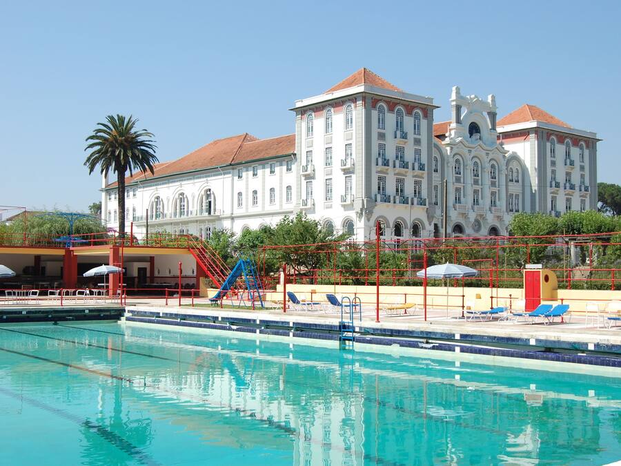 Uma piscina com espreguiçadeiras e guarda-sóis, uma palmeira, e um edifício branco grande com telhado vermelho ao fundo.