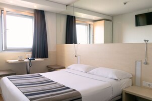 Quarto de hotel com cama, mesa de cabeceira, espelho, televisão na parede, janela com cortinas e zona de duche.
