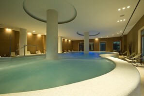 Piscina interna com design curvo, rodeada por espreguiçadeiras e teto moderno com iluminação embutida.