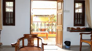Quarto com mesa de madeira, duas cadeiras, um banco e vista para uma varanda com grades e plantas.