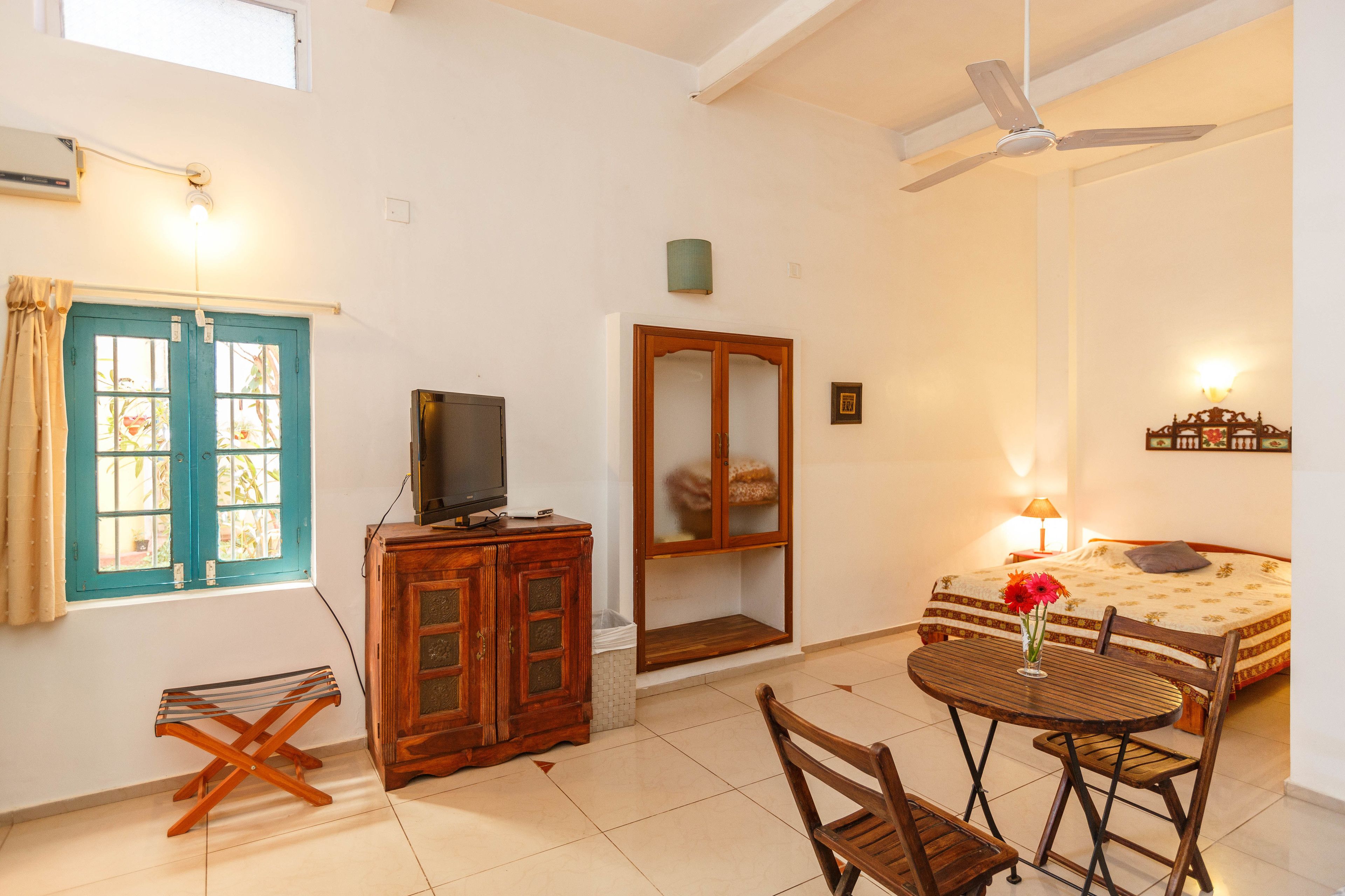 Quarto com cama, televisão, mesa de jantar com cadeiras, armário de madeira e janela com cortinas.