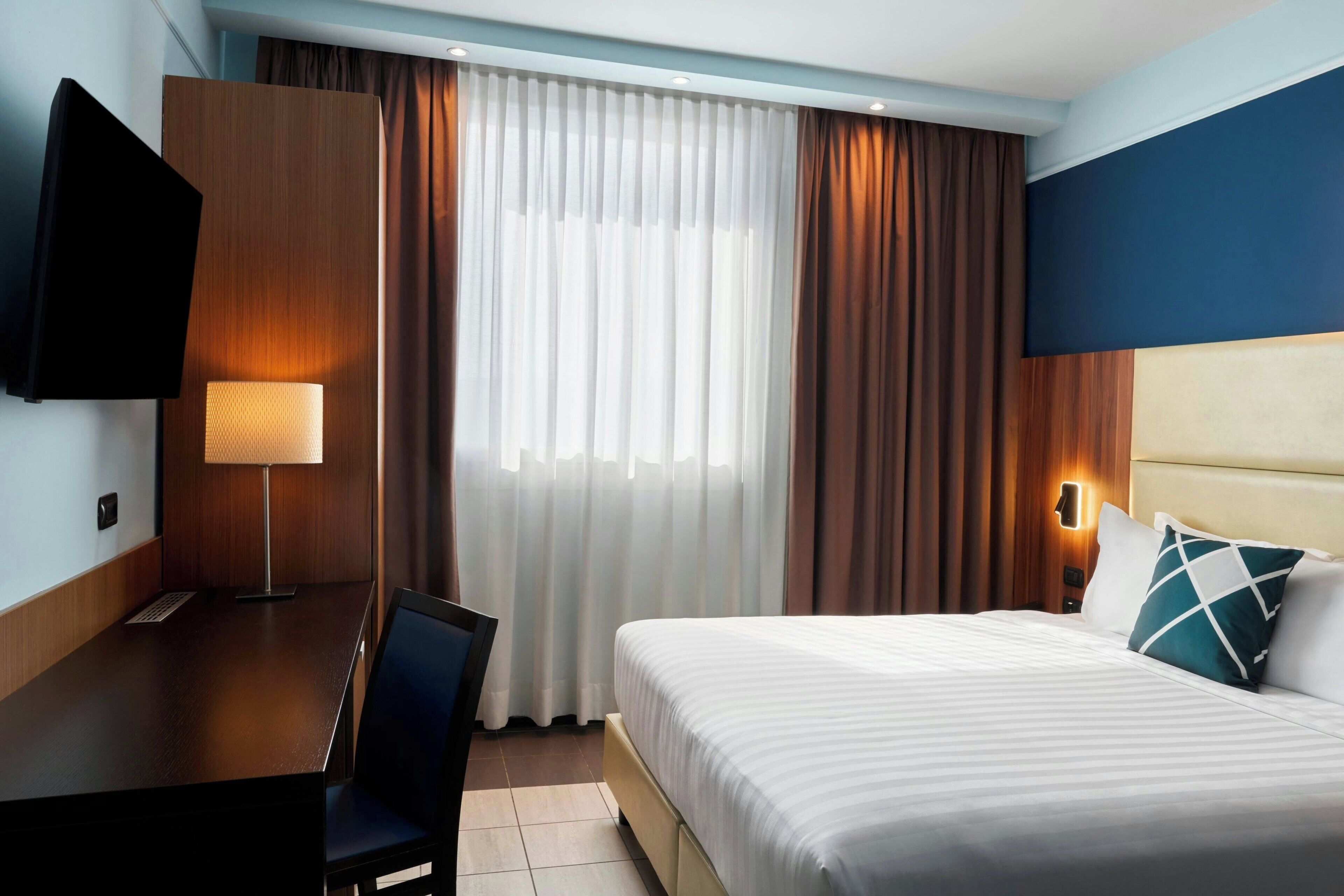 Quarto de hotel com uma cama, uma escrivaninha, uma cadeira, uma televisão e uma janela com cortinas.