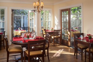 Uma sala de jantar com várias mesas e cadeiras de madeira, janelas amplas e um lustre.