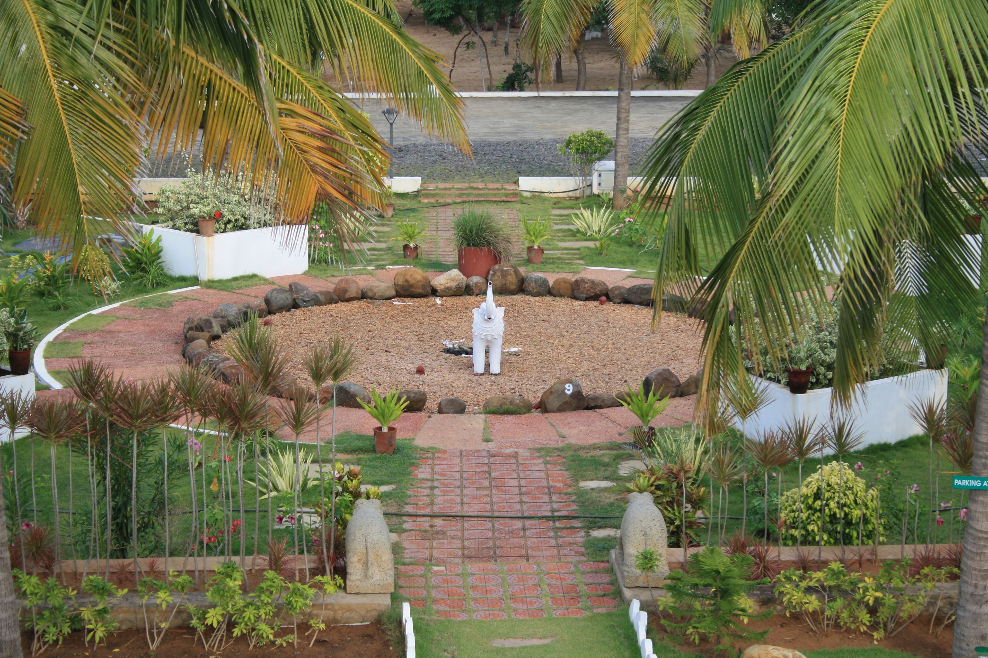 Um jardim circular com uma estátua branca no centro, rodeado por palmeiras e diversas plantas.